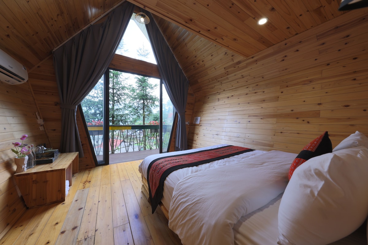 Quarto de cabana de madeira com uma cama, uma mesinha de cabeceira, uma janela com cortinas e vista para árvores.