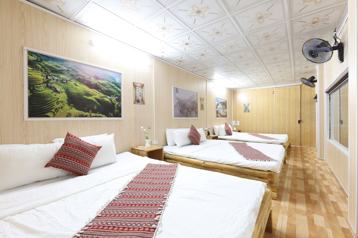 Quarto de hotel com duas camas, uma pintura de paisagem grande e um ventilador de teto.