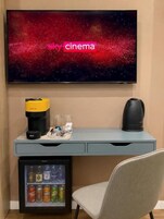 Uma televisão exibindo 'sky cinema', uma cafeteira, um mini-frigobar com bebidas e uma cadeira.