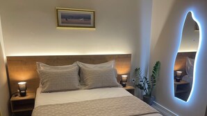 Quarto moderno com uma cama grande, duas luminárias de cabeceira, mesa de cabeceira, planta em vaso, espelho fixado na parede e quadro sobre a cama.