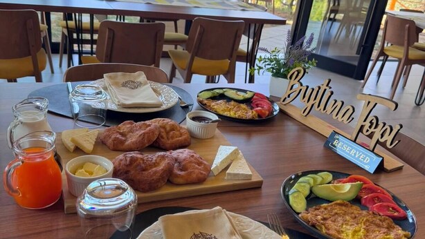 Uma mesa de jantar com diversas opções de pequeno-almoço, incluindo ovos, pão e fruta, numa sala com amplas janelas e vista para o verde.