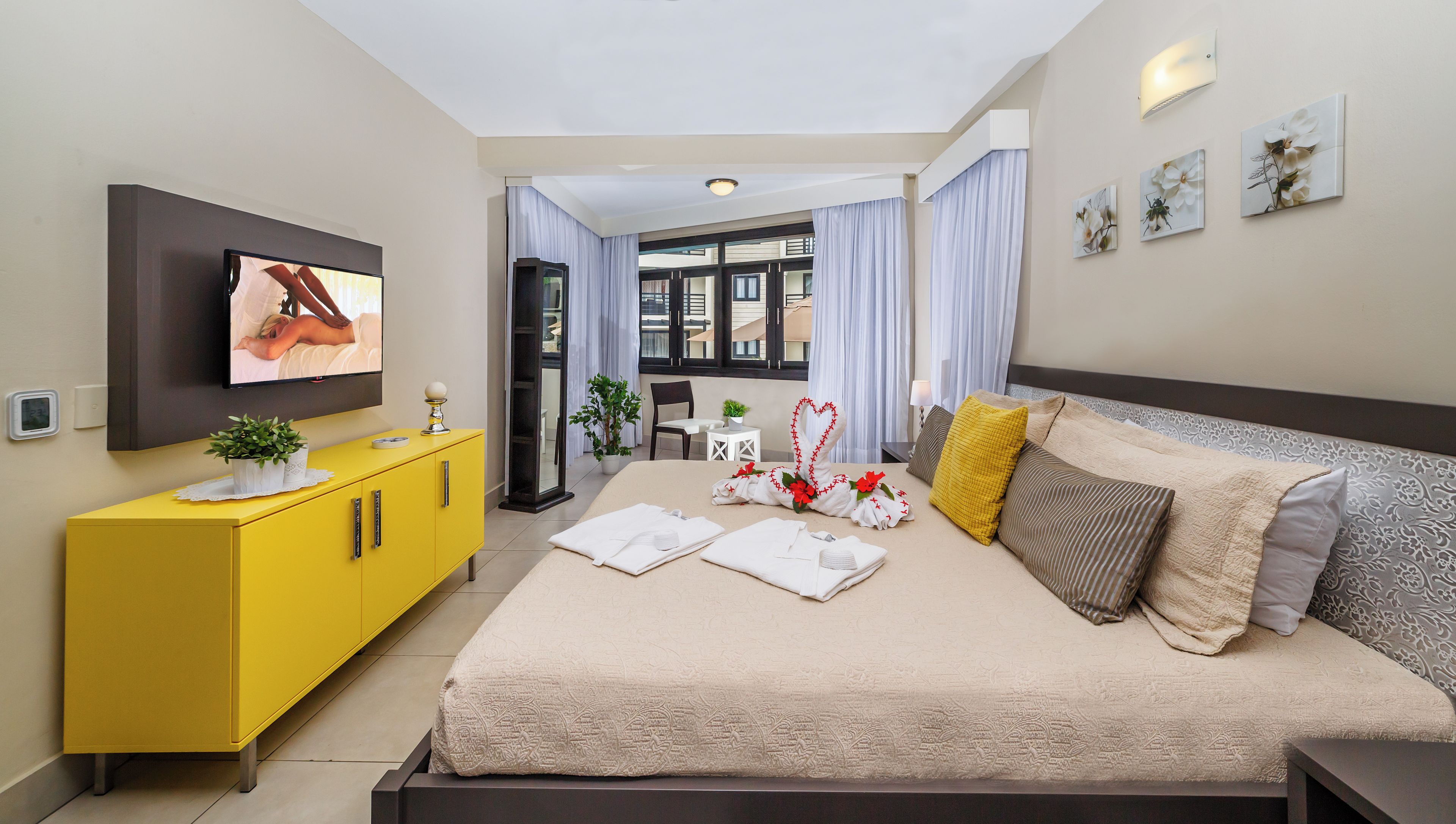 Quarto de hotel com uma cama, televisão, cómoda amarela, mesa pequena com uma planta e vista para o exterior.