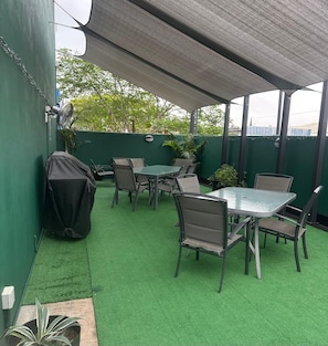 Um pátio externo com mesa e cadeiras, churrasqueira e área coberta com ventilador.