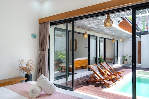 Quarto moderno com uma cama, porta de vidro que dá para uma área de piscina e duas cadeiras de descanso.