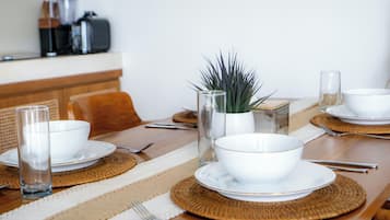 Mesa de jantar posta com pratos, tigelas e copos brancos, acompanhada por um jogo americano tecido e uma pequena planta em vaso.