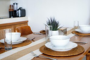 Mesa de jantar posta com pratos, tigelas e copos brancos, acompanhada por um jogo americano tecido e uma pequena planta em vaso.