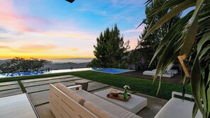 Uma área de estar moderna ao ar livre com piscina, vista para as montanhas e um pôr do sol.