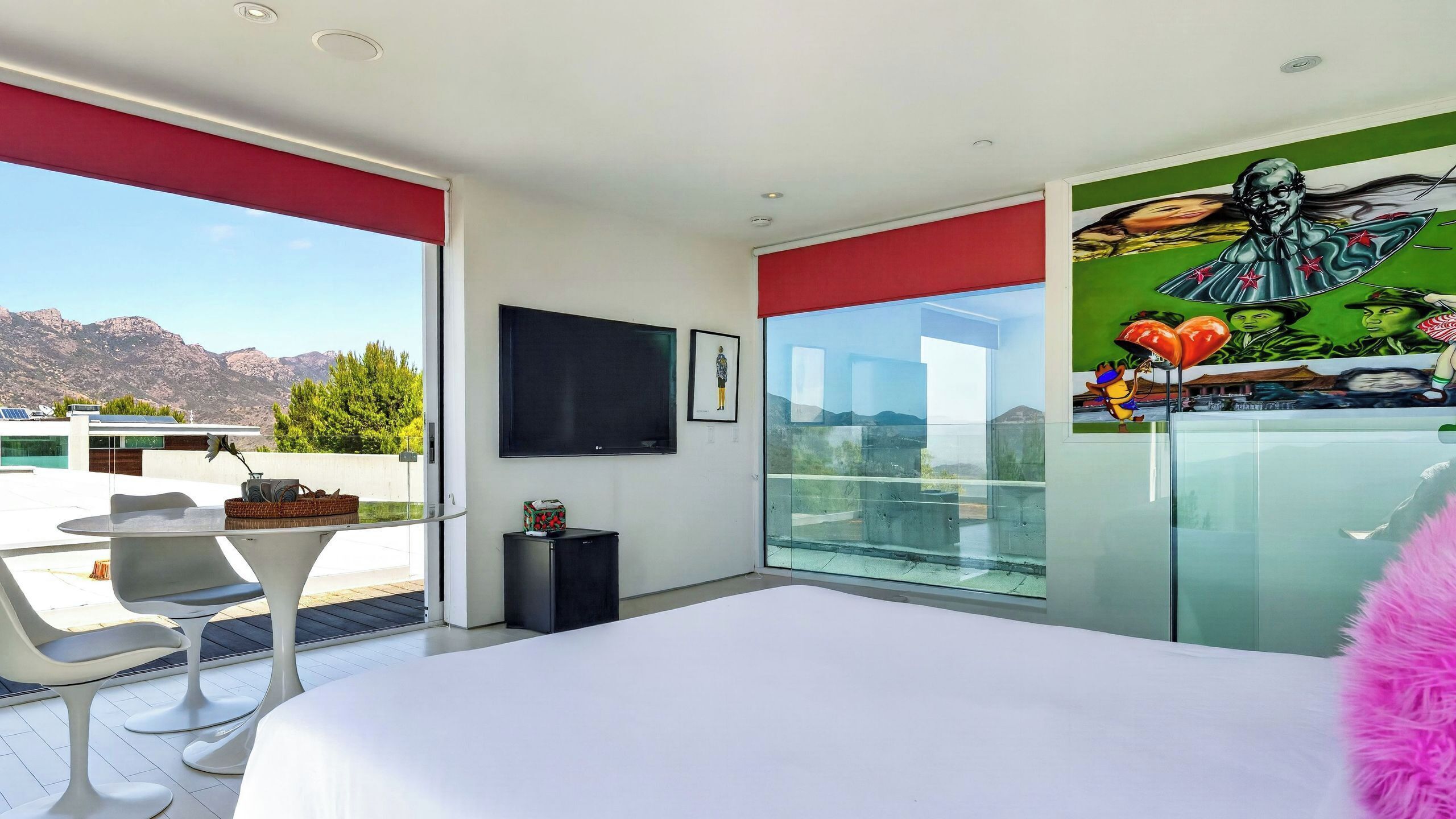 Quarto de hotel moderno com uma cama grande, uma televisão de tela plana, uma área de jantar com mesa e cadeiras, e uma varanda com vista para as montanhas.