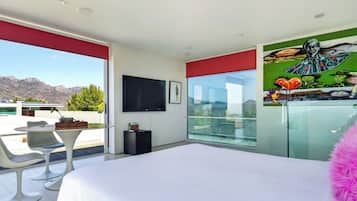 Quarto de hotel moderno com uma cama grande, uma televisão de tela plana, uma área de jantar com mesa e cadeiras, e uma varanda com vista para as montanhas.
