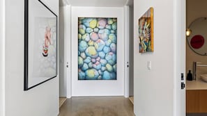 Um corredor com uma porta, um quadro emoldurado à esquerda e uma parede com uma pintura abstrata colorida.