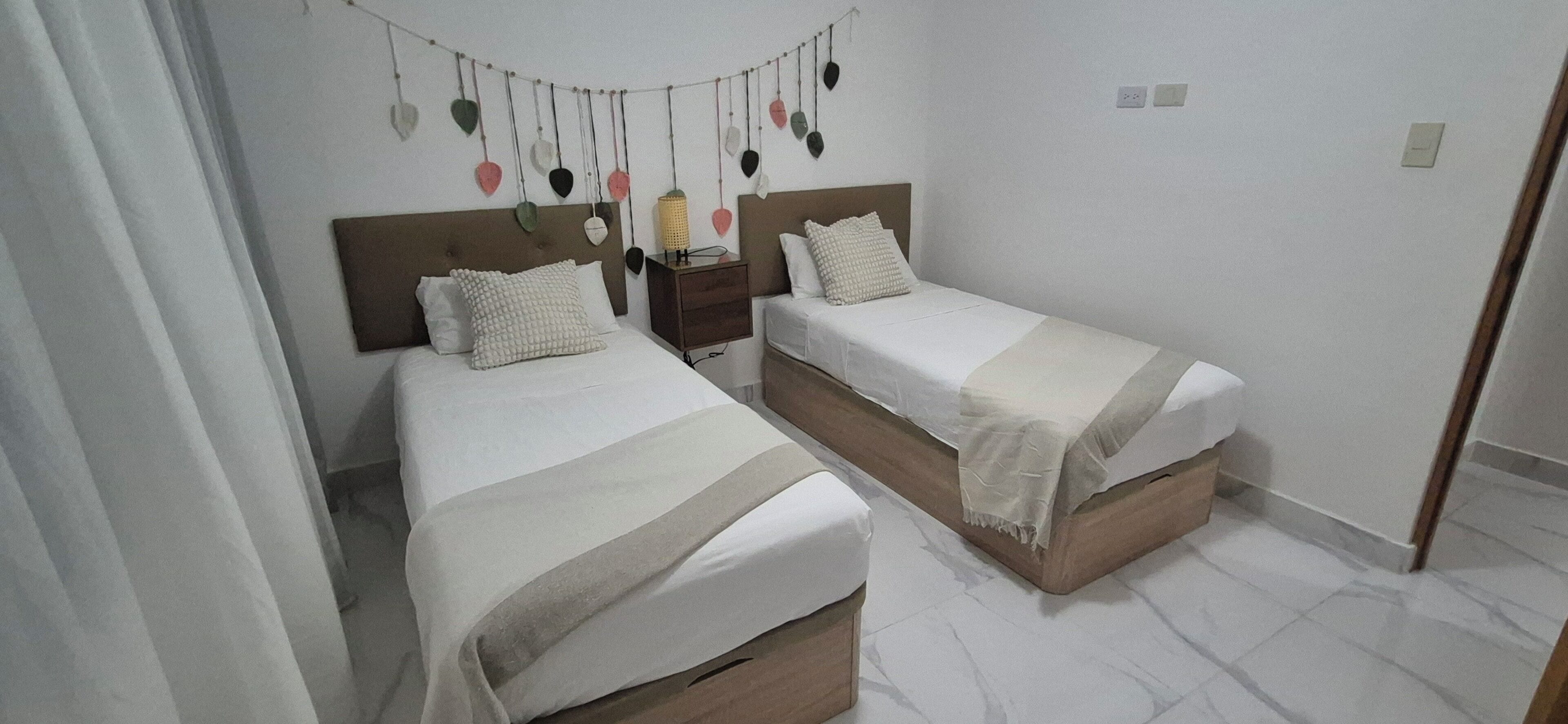 Quarto de hotel com duas camas de solteiro, mesa de cabeceira, abajur e um objeto decorativo fixado na parede.
