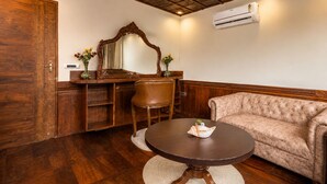 Quarto com teto de madeira, uma mesa redonda com concha decorativa, sofá acolchoado, escrivaninha de madeira com espelho e unidade de ar condicionado.