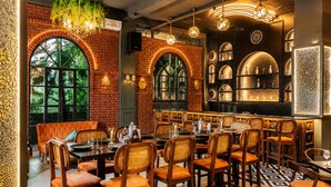 Interior de restaurante com paredes de tijolo, janelas em arco e área de bar com prateleiras de garrafas.