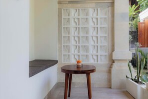 Uma pequena mesa de madeira com um objeto decorativo, parede branca com padrão geométrico e uma planta em vaso branco.