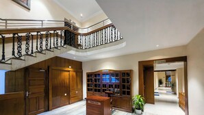 Um saguão de hotel com piso de mármore, balcão de recepção em madeira, escada com grades ornamentadas e uma estante de livros.