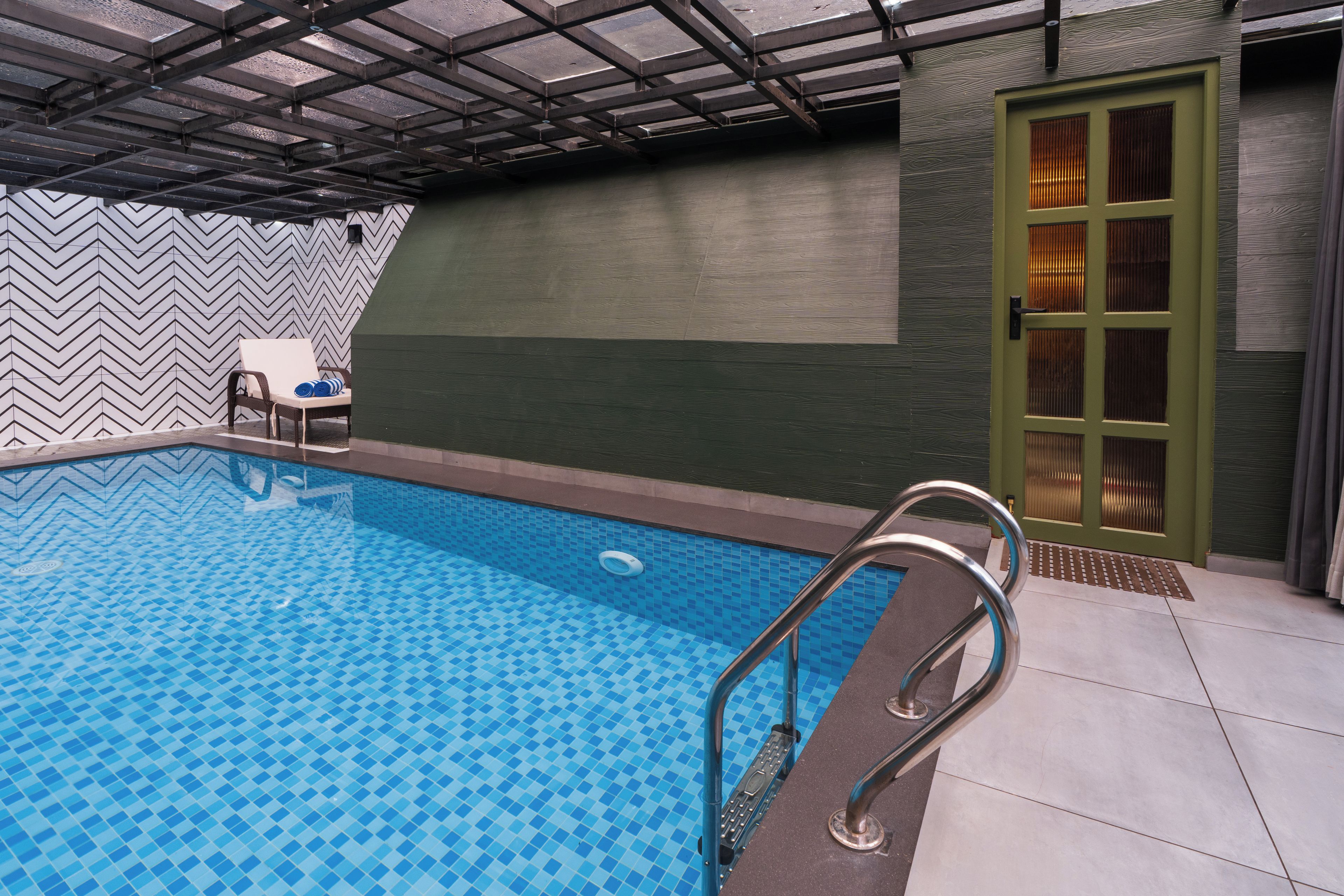 Piscina interna com parede em padrão geométrico, porta com janela e cadeira com toalhas.