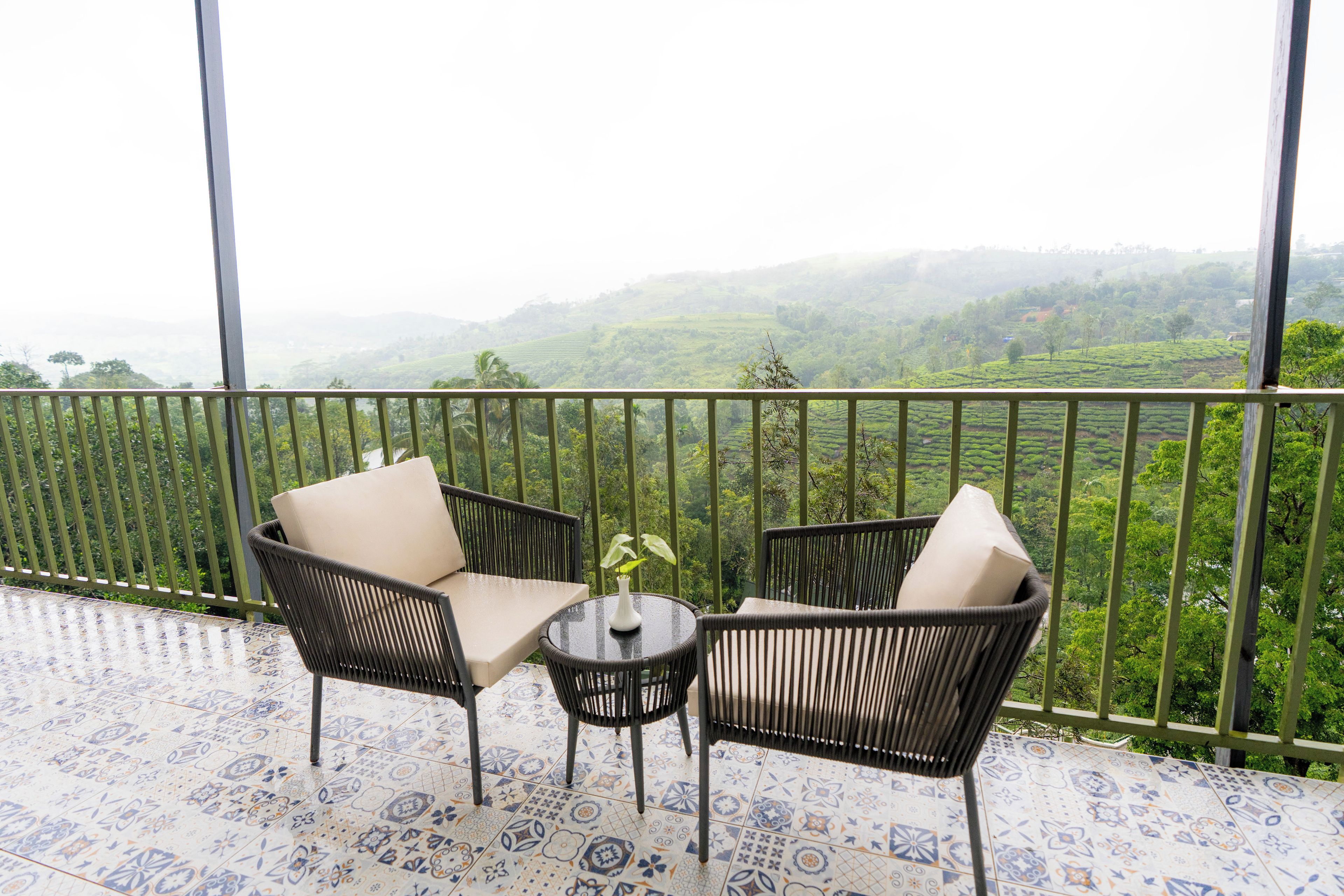 Uma varanda com duas cadeiras de vime e uma pequena mesa, com vista para uma paisagem verde exuberante.