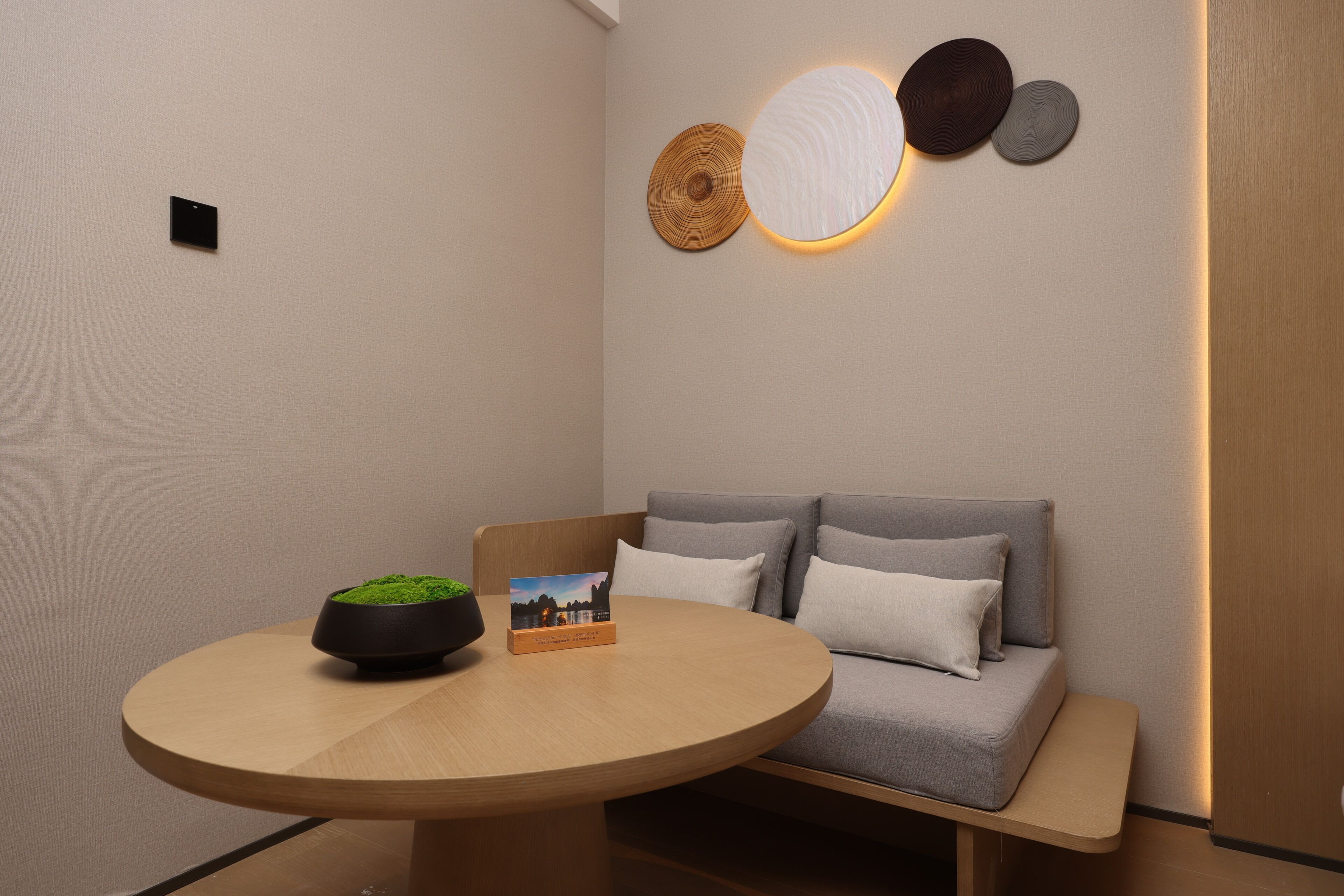 Quarto moderno com uma mesa redonda de madeira, um sofá cinza com almofadas e um lustre fixado na parede.