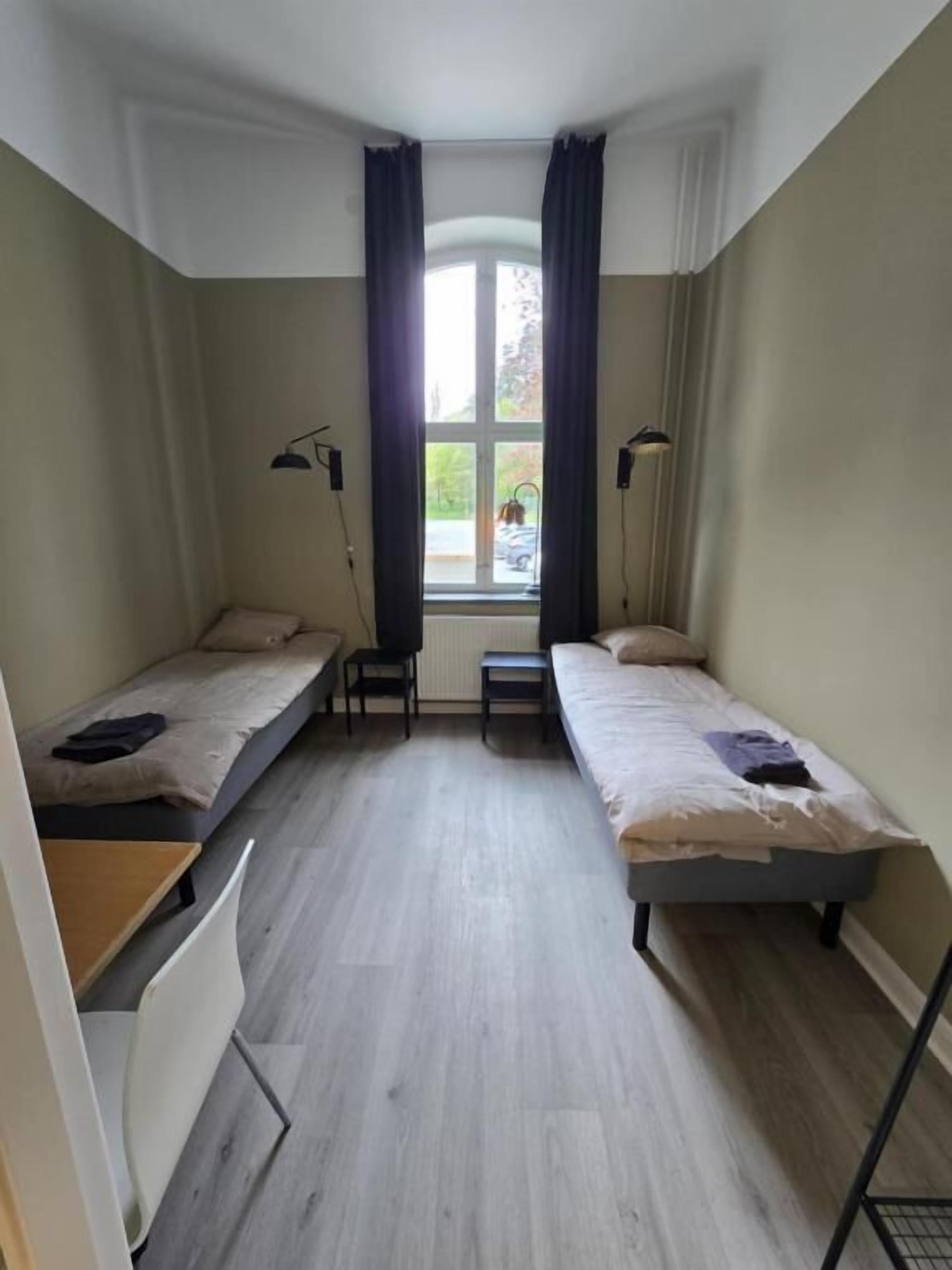 Quarto com duas camas de solteiro, uma mesinha, uma cadeira e uma janela com cortinas.