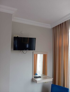 Quarto de hotel com televisão na parede, espelho sobre uma escrivaninha e janela com cortinas.