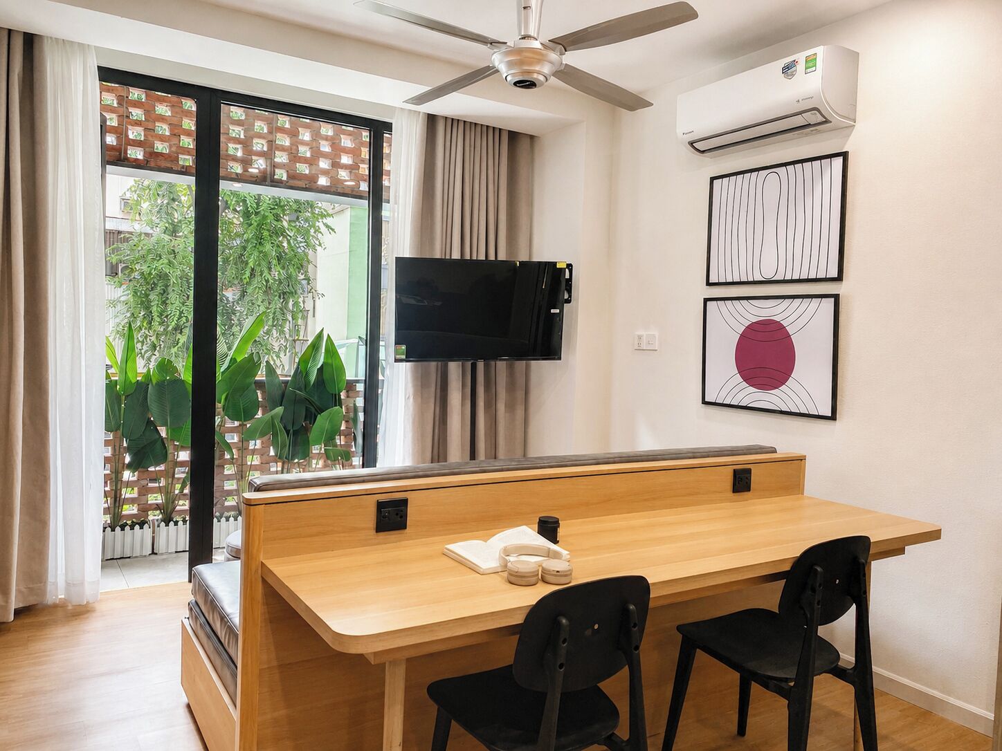 Quarto moderno com escrivaninha de madeira, duas cadeiras pretas, ventilador de teto, ar condicionado e TV fixada na parede.