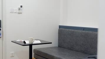 Quarto pequeno e moderno, com um sofá cinza, uma mesa preta com uma caneca branca e uma tomada na parede.