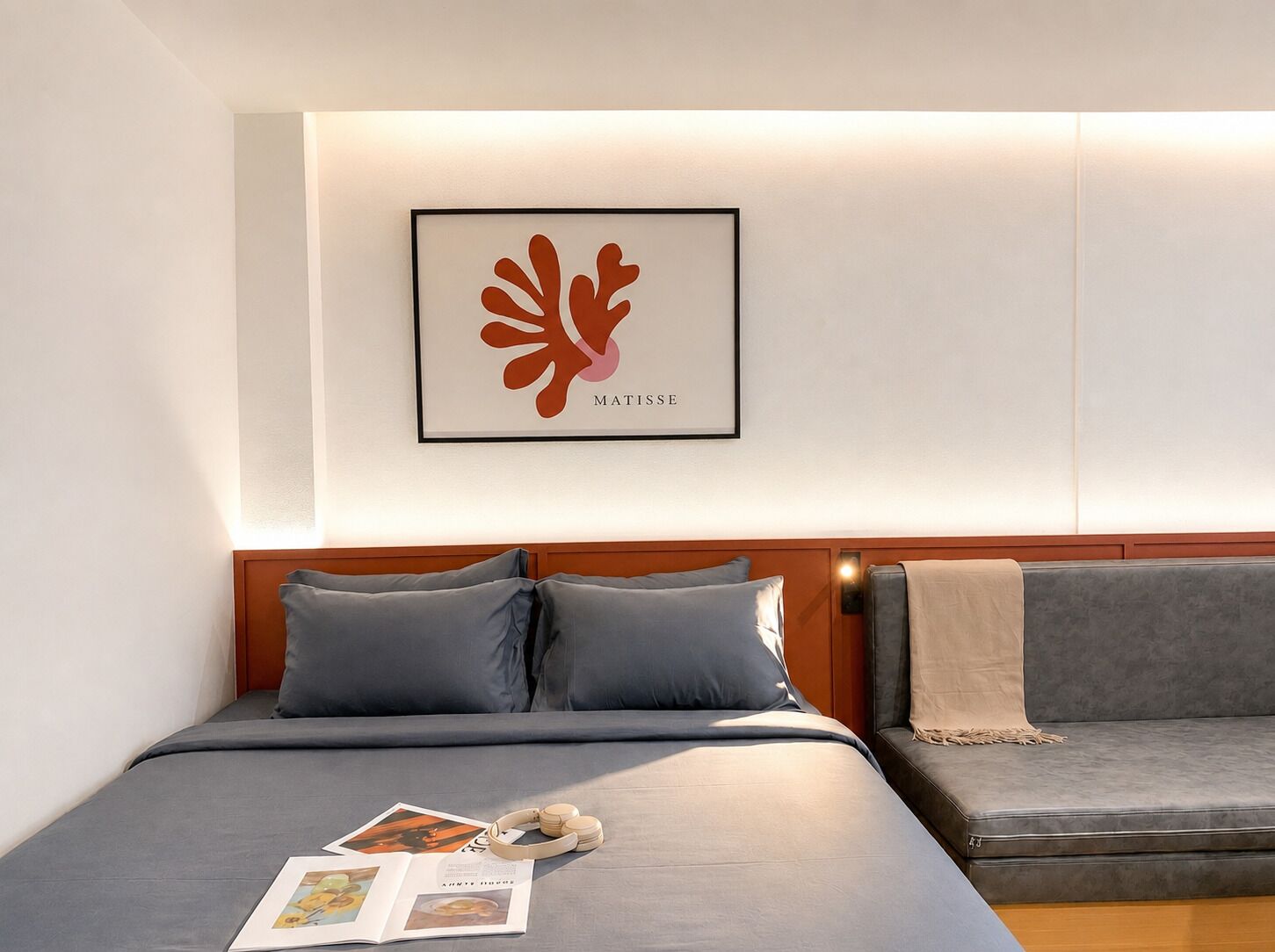 Cama bem arrumada com colchão e almofadas cinzas, uma obra de arte emoldurada na parede e um sofá cinza com um cobertor.