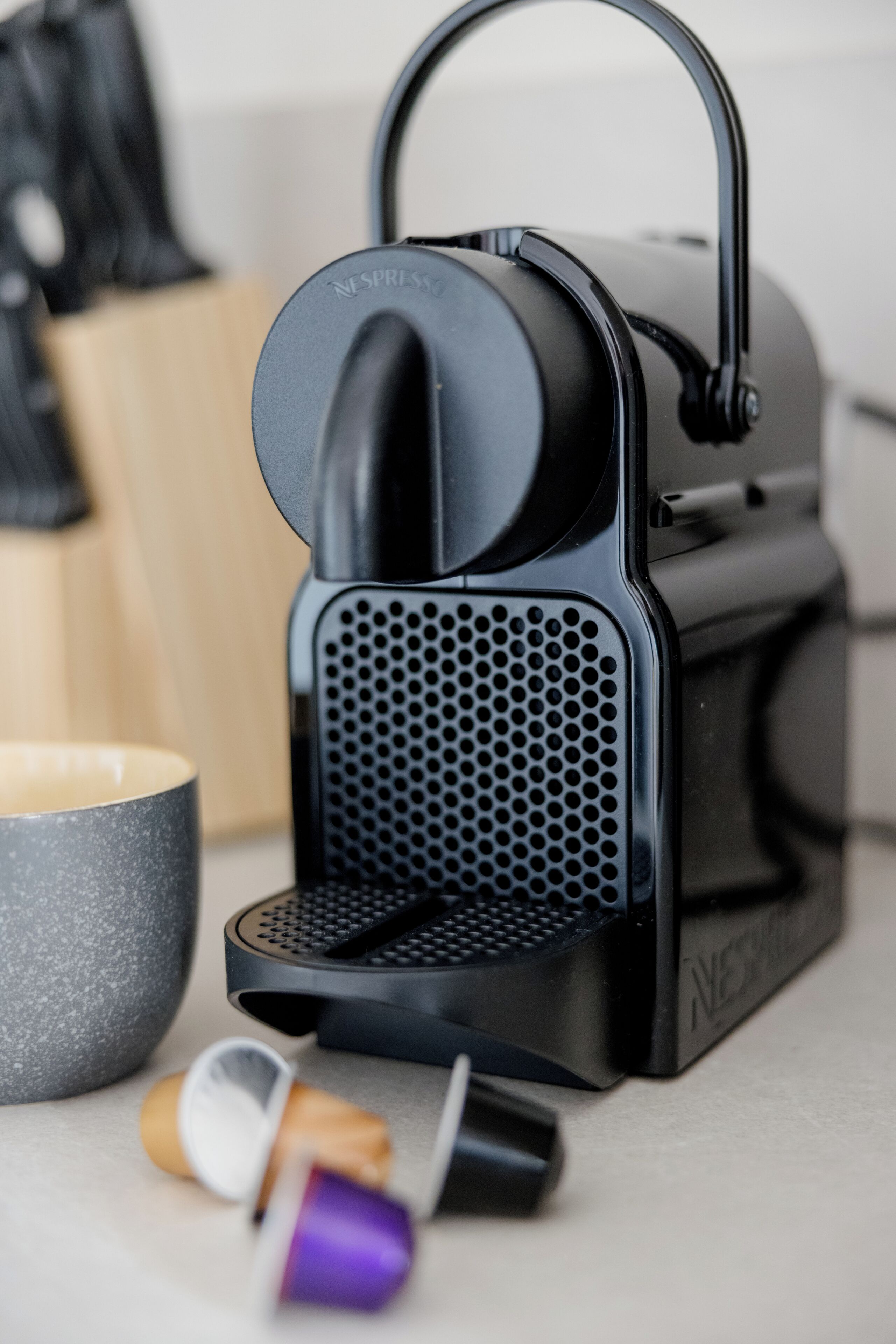 Uma máquina de café Nespresso preta com um porta-xícaras, uma xícara de café e algumas cápsulas de café em uma bancada.