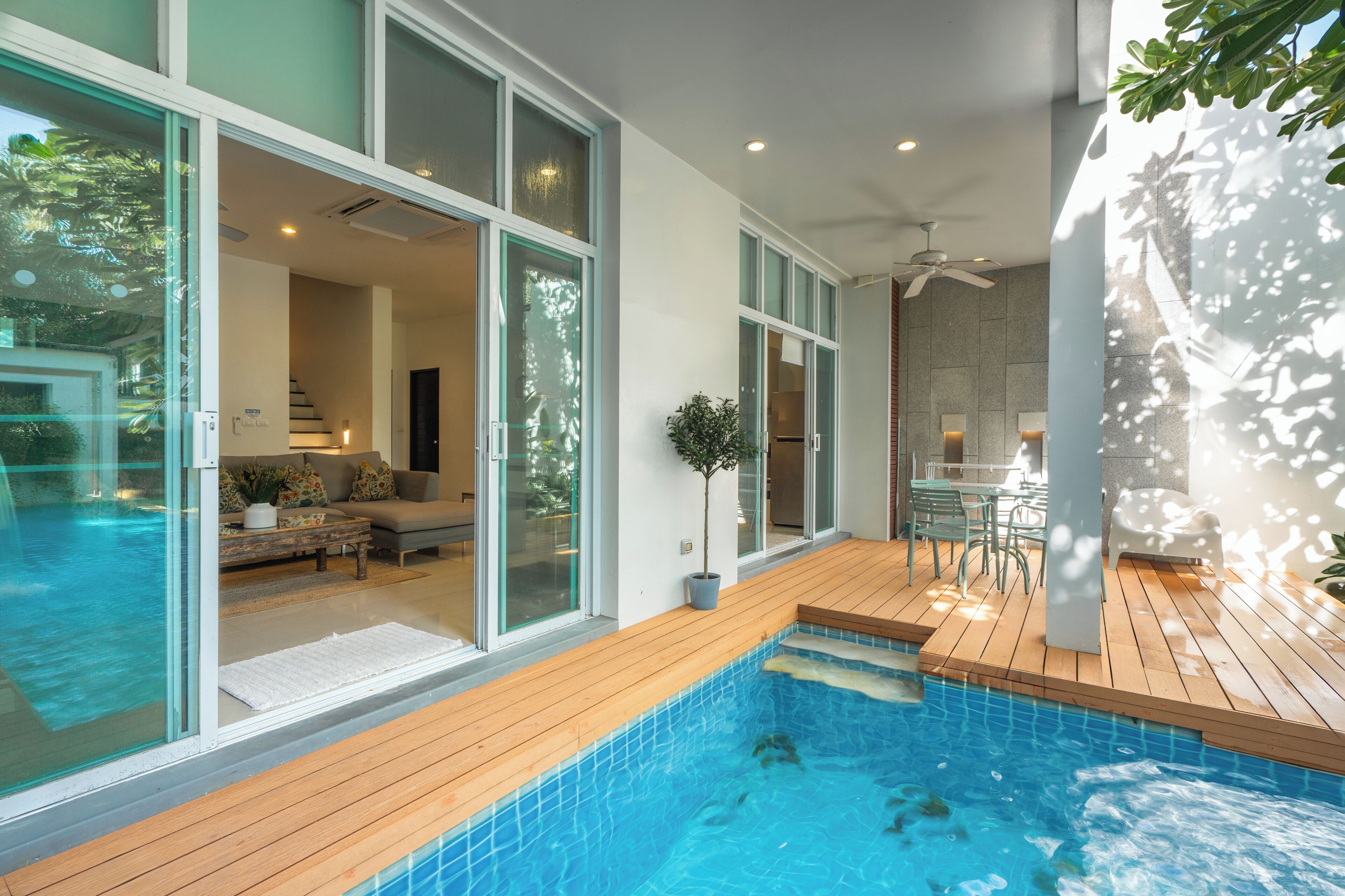 Piscina interior moderna com água azul cristalina, ladeada por um deck de madeira e portas de vidro que dão para uma sala de estar com sofá e mesa de jantar.