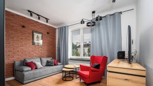 Uma sala de estar moderna com parede de tijolos, um sofá cinza, uma poltrona vermelha, uma escrivaninha de madeira e uma janela com cortinas.