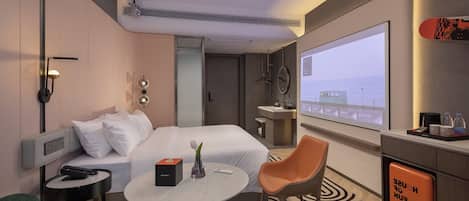 Quarto de hotel com cama, mesa de cabeceira, cadeira, escrivaninha e uma grande tela de TV.