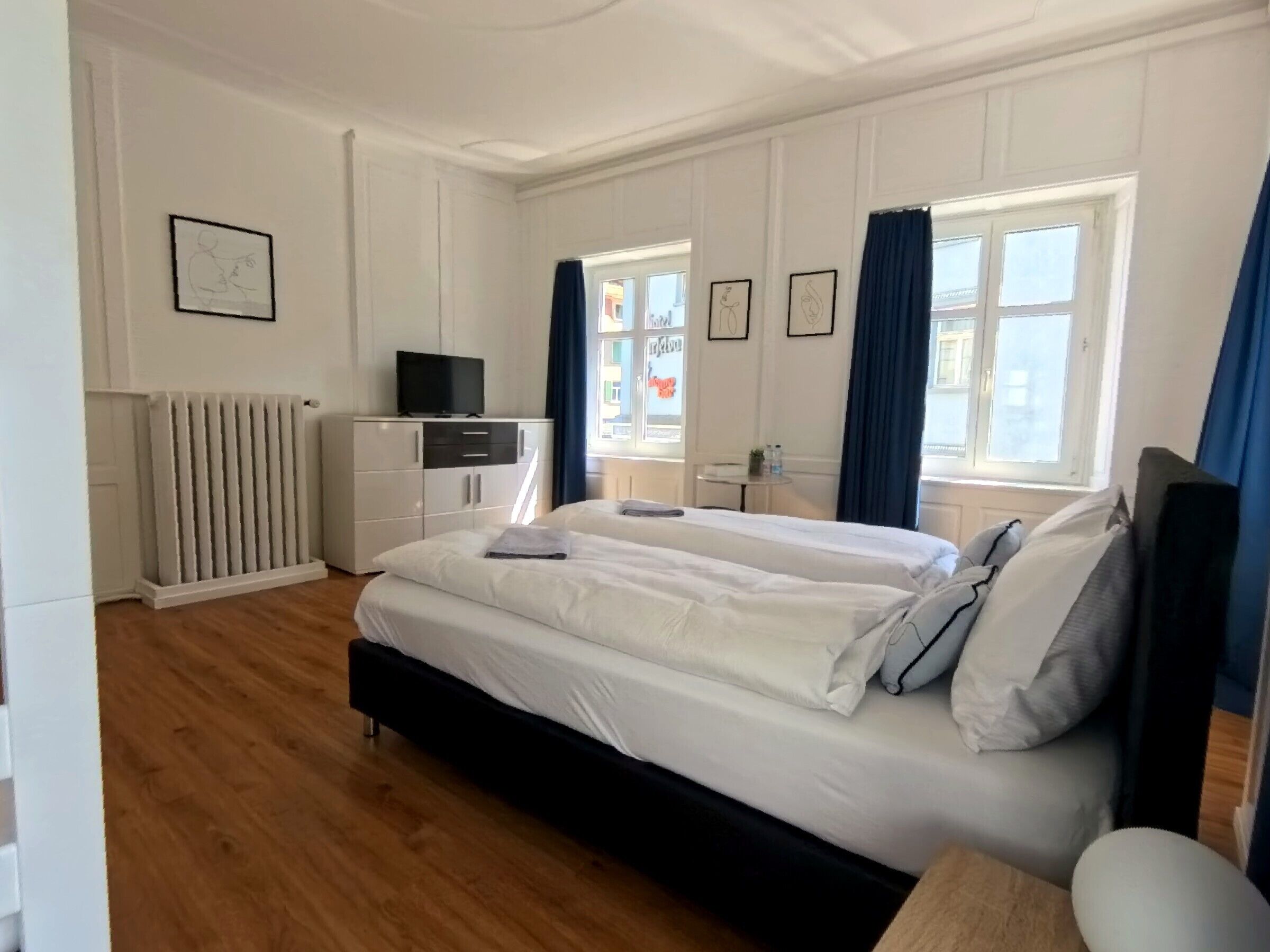 Quarto de hotel com cama de casal, mesa de cabeceira, cômoda, radiador e janela com cortinas.