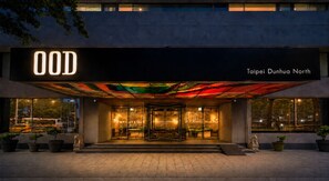 Entrada de hotel com fachada de vidro, área de estar ao ar livre e teto colorido.