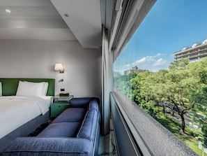 Quarto de hotel com cama, mesa de cabeceira, luminária e vista para a paisagem urbana através de uma janela grande.