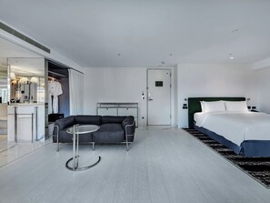 Quarto de hotel moderno com uma cama, área de estar, toucador com espelho e porta para outro espaço.