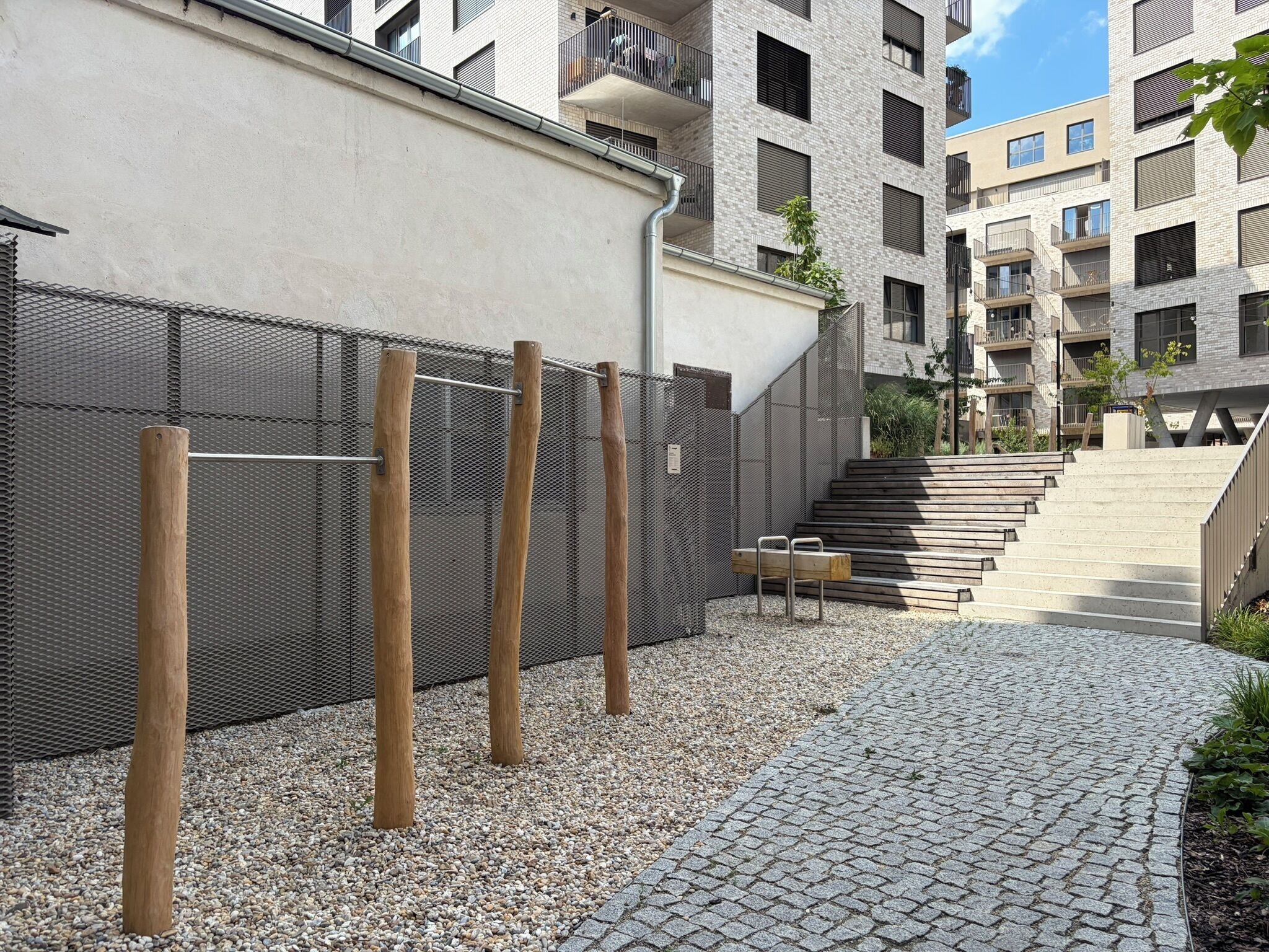 Um pátio com cerca de metal, postes de madeira, um banco e uma escadaria que leva a um prédio de apartamentos.