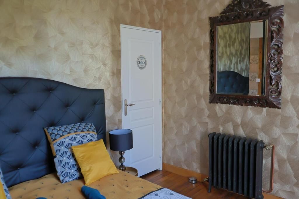 Quarto com cabeceira estofada, almofadas estampadas, luminária de cabeceira, radiador e espelho com moldura ornamentada.