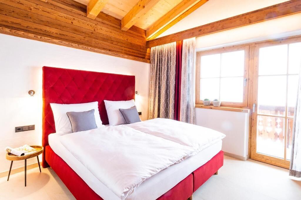 Quarto de hotel com uma cama grande, vigas de madeira no teto, uma janela com cortinas e uma pequena mesa de cabeceira.