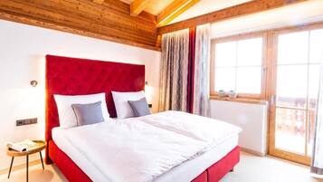 Quarto de hotel com uma cama grande, vigas de madeira no teto, uma janela com cortinas e uma pequena mesa de cabeceira.