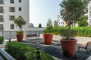 Um jardim no terraço com plantas em vasos, um banco e vista para edifícios vizinhos.