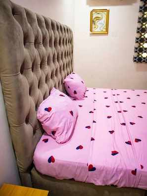 Uma cama com cabeceira estofada, edredom rosa com estampas de corações e uma única almofada rosa.