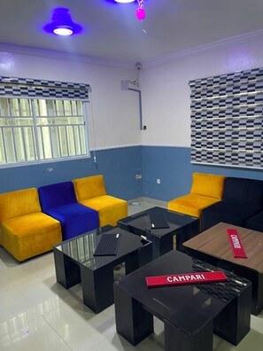 Um quarto com persiana xadrez, sofá azul e amarelo, e uma mesa com um letreiro vermelho.