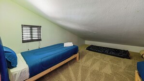 Quarto pequeno para uma pessoa, com beliche, janela com persianas, piso coberto por carpete e tapete preto no chão.