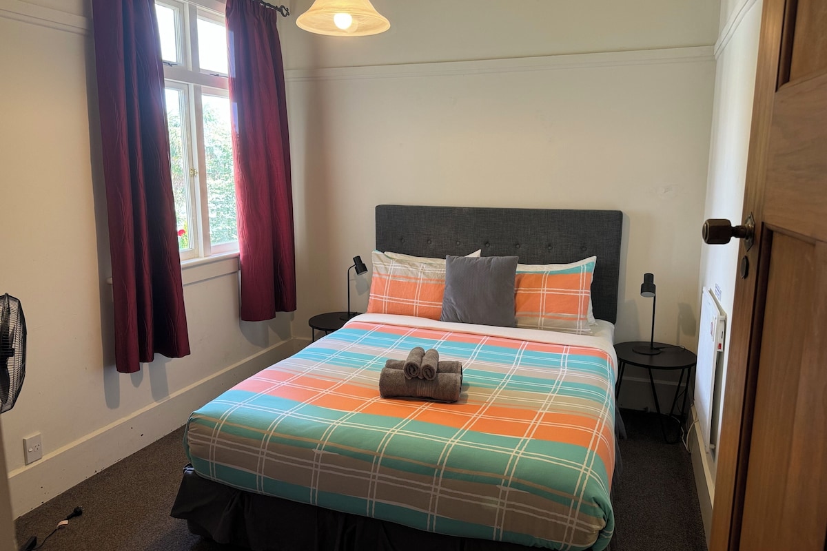 Um quarto com uma cama, uma janela com cortinas, um abajur e um ventilador fixado na parede.