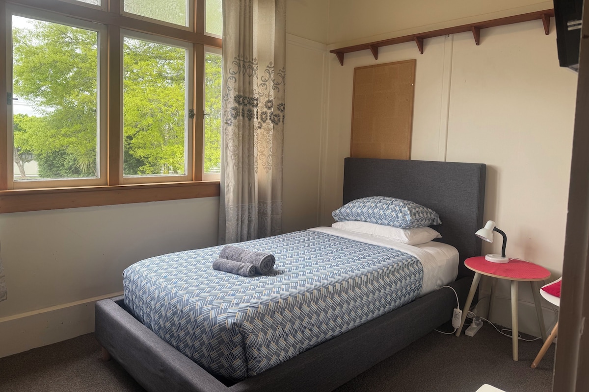 Quarto com cama, janela com cortinas, mesa de cabeceira com abajur e vista para árvores.