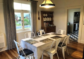 Uma área de refeições com uma mesa posta para quatro pessoas, cadeiras brancas, uma janela com cortinas, um armário de madeira com louças e uma cozinha visível através de uma porta aberta.
