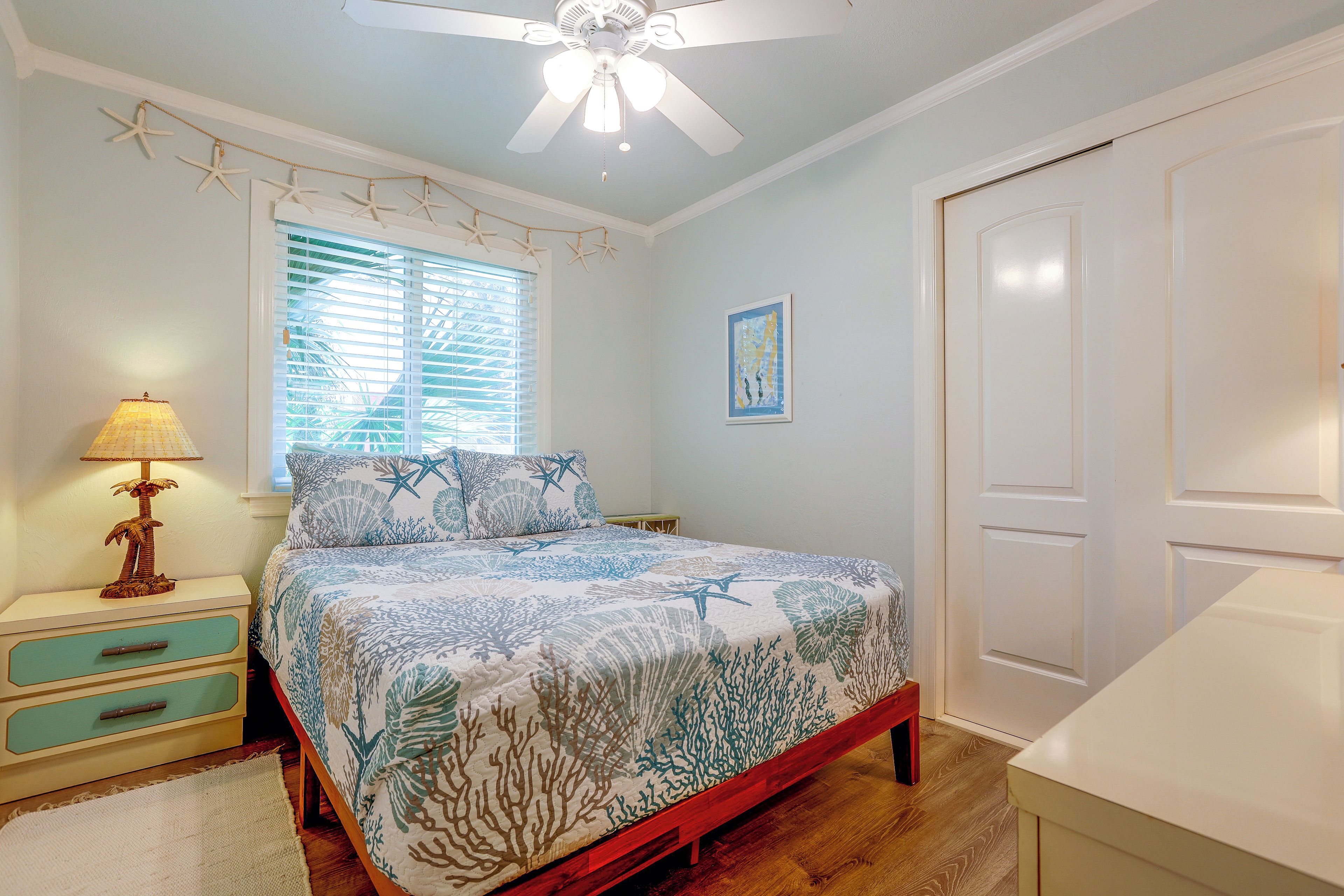 Quarto com cama, mesa de cabeceira, abajur, janela com persianas, ventilador de teto e porta.