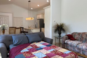 Uma sala de estar com um sofá cinzento, uma poltrona estampada, um cobertor acolchoado, uma área de jantar com um candelabro e uma cozinha com armários brancos.