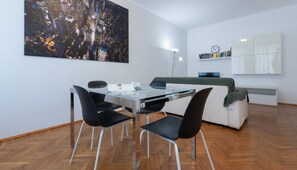 Uma sala de estar moderna com uma mesa de jantar de vidro, cadeiras pretas, um sofá branco e um grande quadro com uma vista urbana na parede.