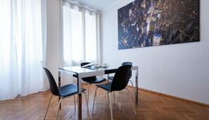 Uma sala de jantar moderna com mesa de vidro, quatro cadeiras e uma pintura de paisagem urbana na parede.